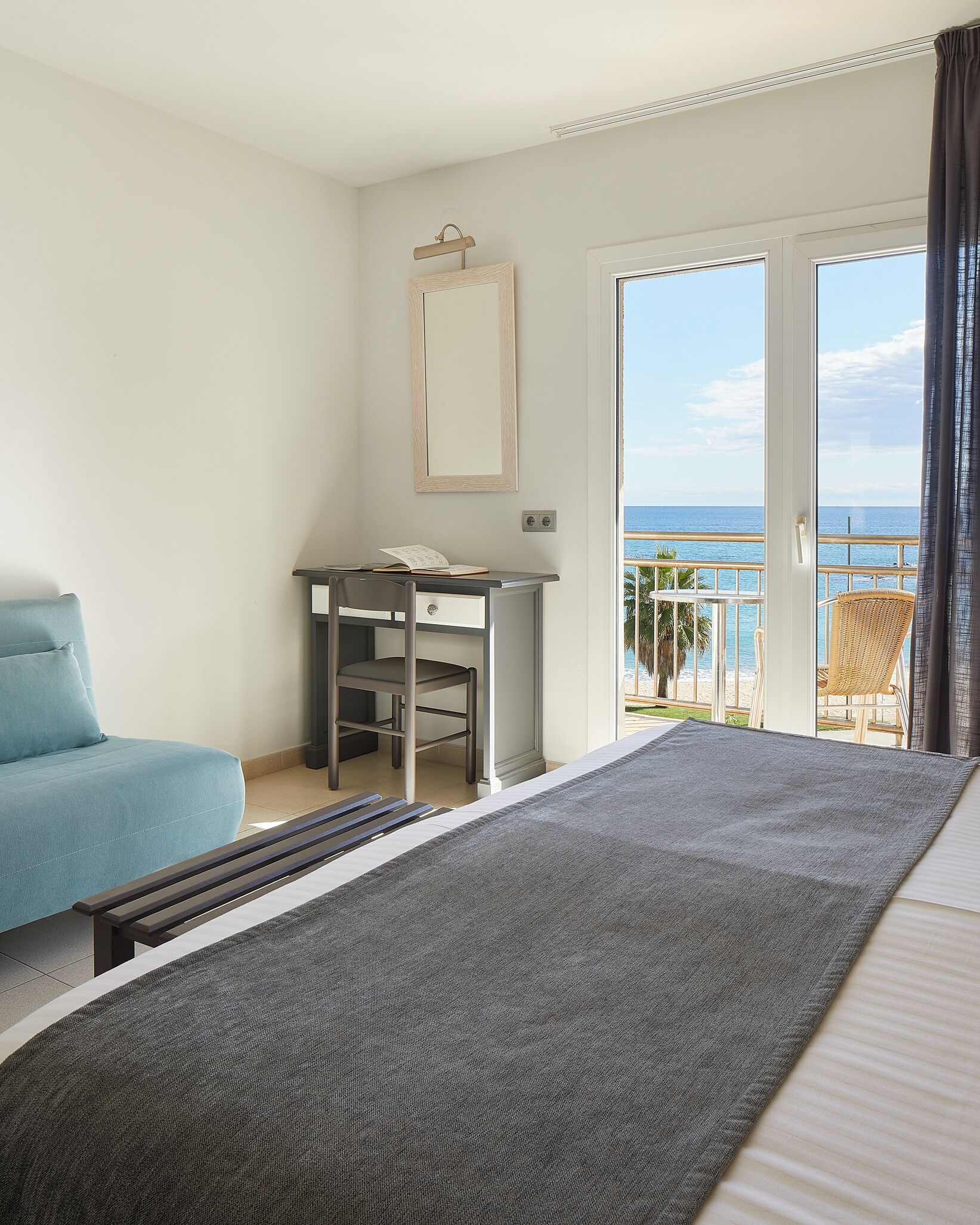 Quarto de hotel com cama, escrivaninha, cadeira e vista para o mar através de amplas janelas.