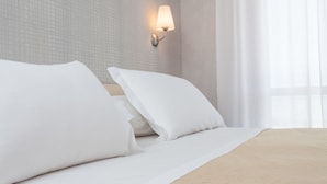 Uma cama bem arrumada com lençóis brancos e cabeceira em tom claro. Uma luminária fixada na parede é visível à direita da cama.