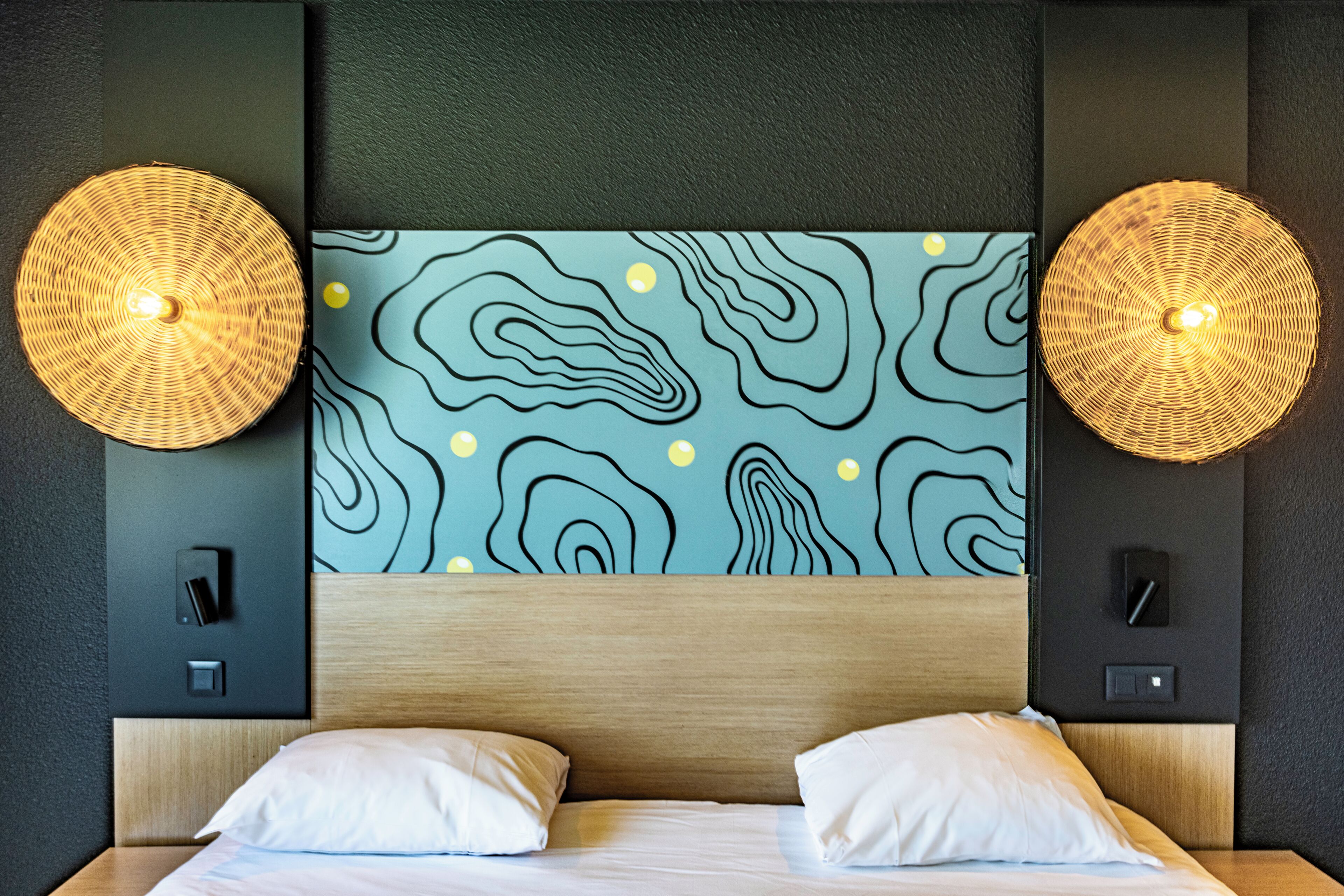 Quarto de hotel moderno com cabeceira de madeira, duas luminárias de apoio e uma parede com arte abstrata.