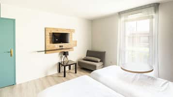Quarto de hotel com uma cama, uma televisão fixada num painel de madeira, uma mesa pequena com uma lâmpada, uma cadeira e uma janela com cortinas.