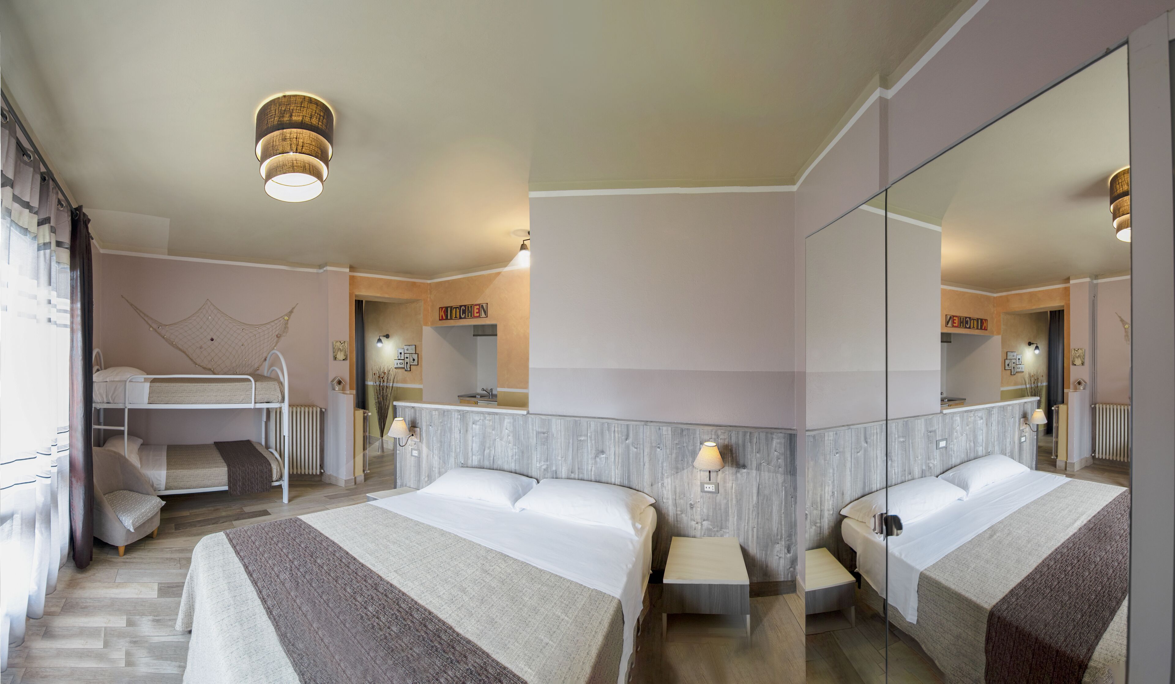 Quarto de hotel com duas camas, uma mesinha, uma cadeira, um espelho e uma área de cozinha.