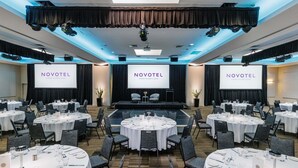 Uma sala de conferências com mesas redondas, palco e uma tela grande exibindo 'NOVOTEL SYDNEY OLYMPIC PARK'.