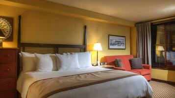Quarto de hotel com uma cama grande, escrivaninha, cadeira, cômoda, abajur e janela com persianas.