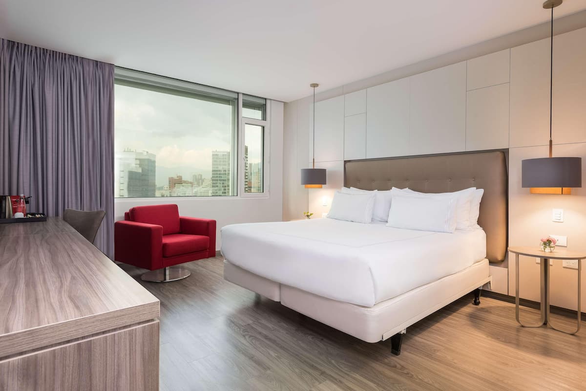 Quarto de hotel com cama, escrivaninha, uma cadeira vermelha e vista para a cidade.