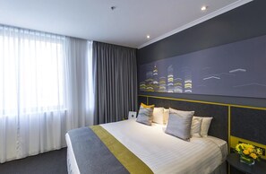 Quarto de hotel com uma cama grande, mesa de cabeceira com um vaso de flores e uma pintura de paisagem urbana na parede.