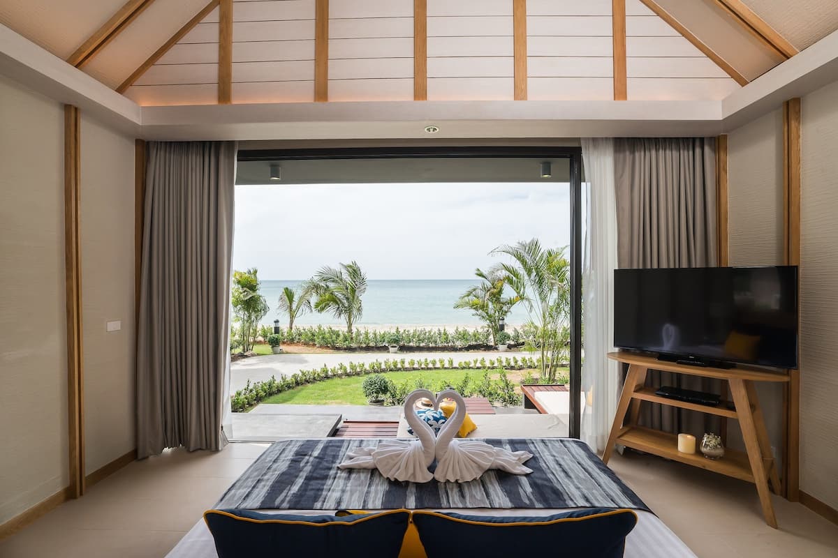 Quarto com uma grande janela, uma cama, uma TV sobre um suporte de madeira e vista para a praia.