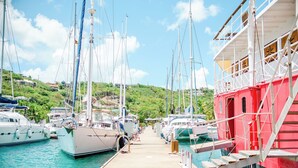 Uma marina com vários barcos atracados, um edifício vermelho com escadas e uma encosta ao fundo.
