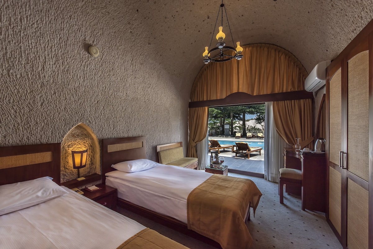 Quarto de hotel com duas camas, cabeceira de madeira, mesa de cabeceira com abajur, vista para uma piscina ao ar livre e área de estar com cadeiras e mesa.