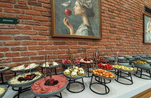 Disposição de buffet com diversos alimentos em bandejas, em frente a uma parede de tijolos com um quadro emoldurado.