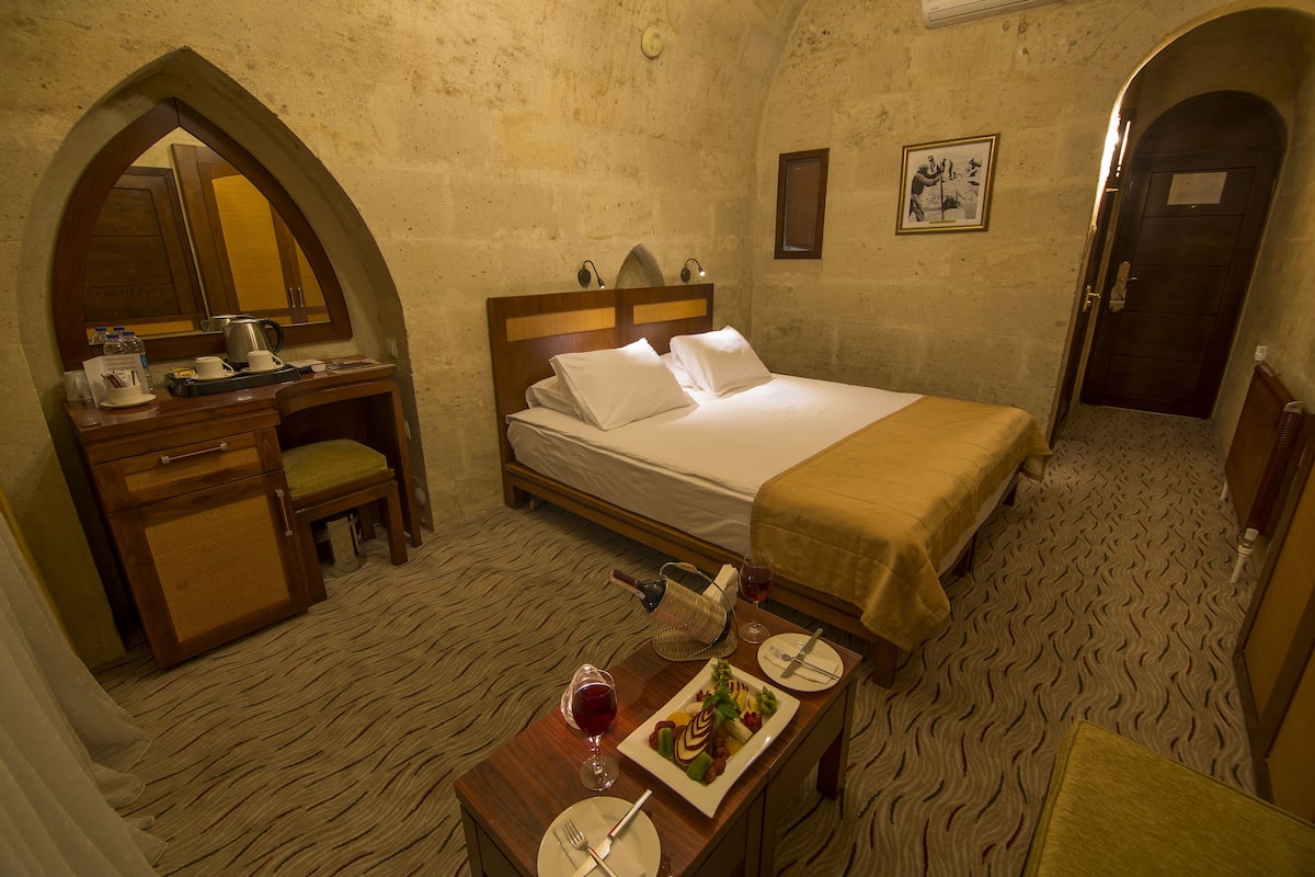 Quarto de hotel com uma cama, uma escrivaninha com uma cafeteira, uma mesa de jantar com um prato de comida e vinho, e uma porta em arco.