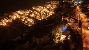 Vista aérea noturna de uma propriedade costeira com formações rochosas iluminadas, piscina e estrada que leva a um edifício.
