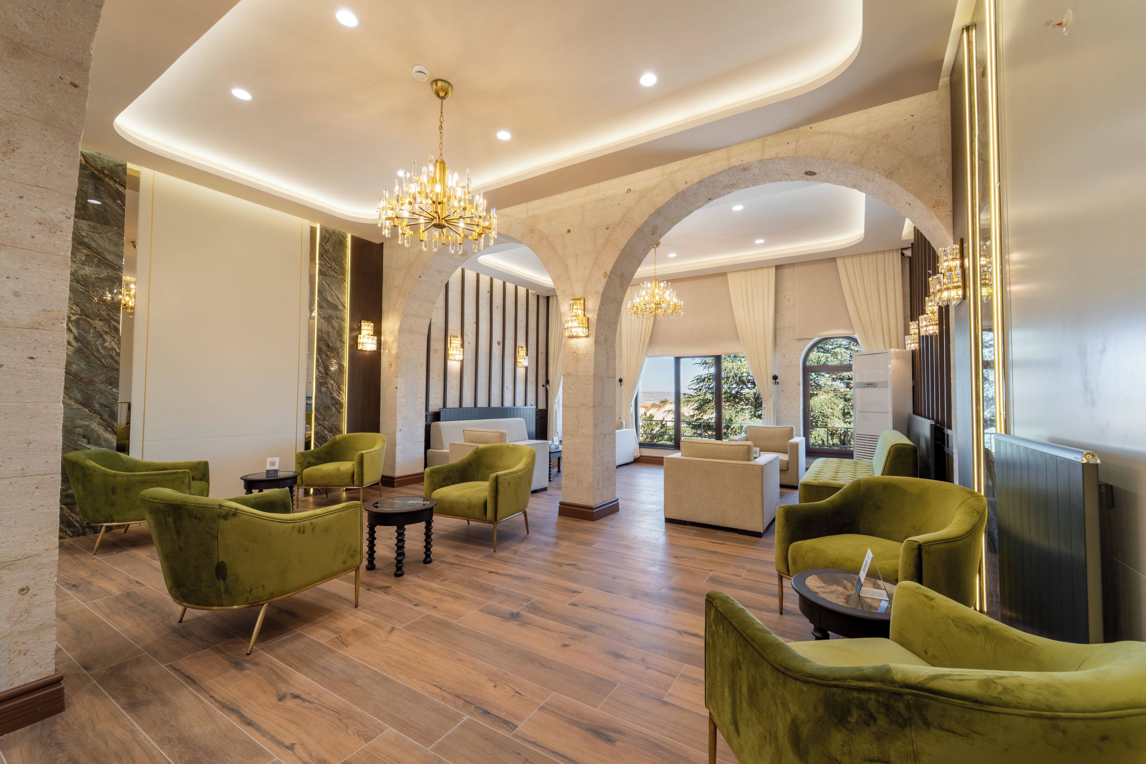Um hall amplo com poltronas estofadas em verde, um lustre imponente e vista para uma varanda através de uma porta arqueada.