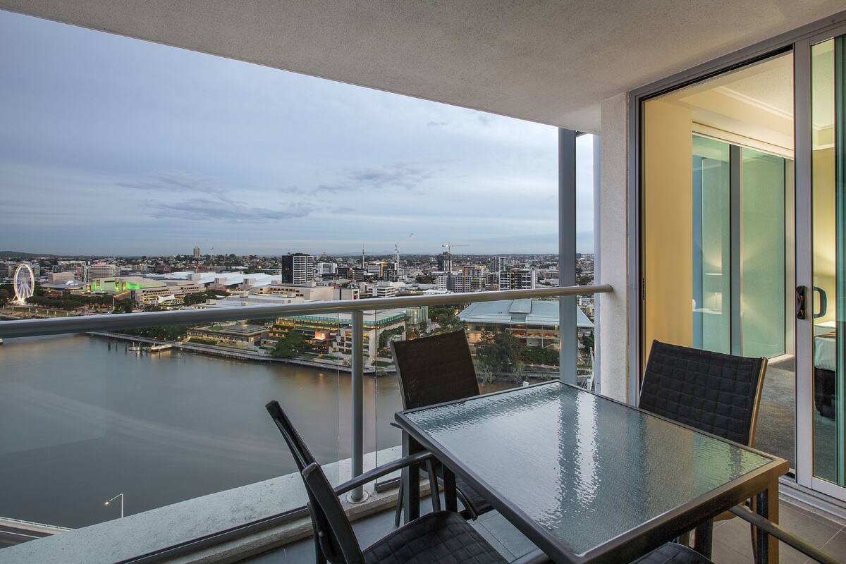 Uma varanda com mesa e cadeiras de vidro, com vista para uma paisagem urbana que inclui um rio e uma roda gigante.