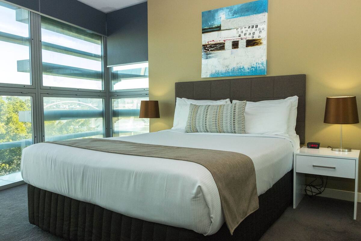 Quarto de hotel moderno com uma cama grande, duas mesas de cabeceira e uma janela com vista para uma ponte.