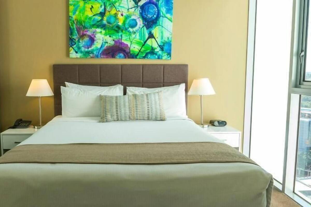 Quarto de hotel com uma cama grande, duas luminárias de cabeceira, um telefone e uma pintura abstrata colorida na parede.