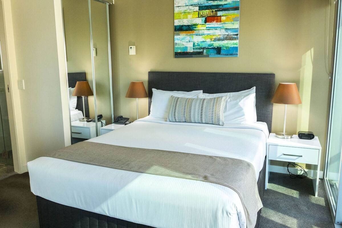 Quarto de hotel com uma cama grande, duas mesas de cabeceira, um criado-mudo, um quadro na parede e uma janela.