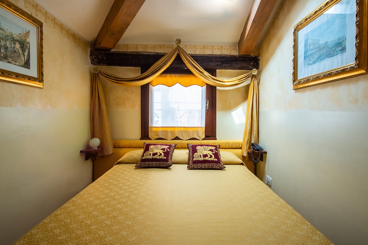 Quarto com cama, duas almofadas decorativas, janela com cortinas e quadros nas paredes.