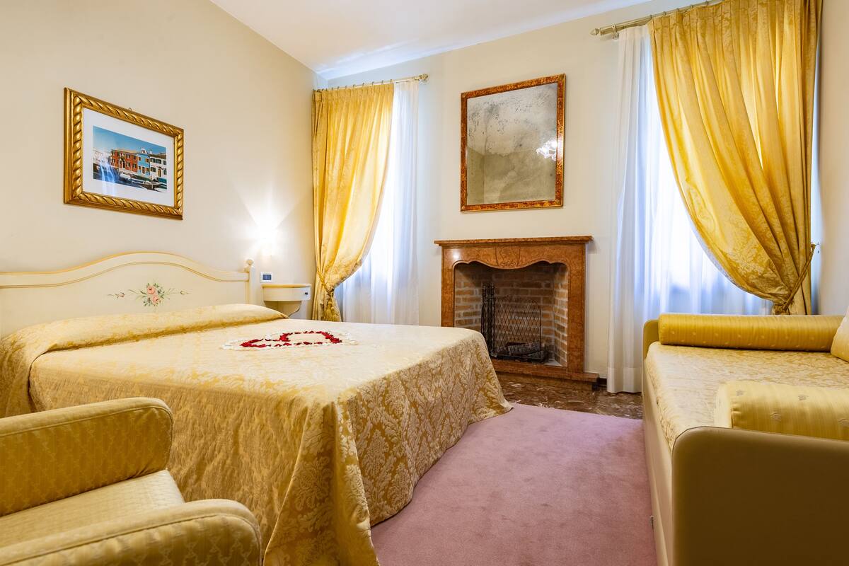 Quarto de hotel com uma cama de casal, duas camas simples, lareira e um espelho grande na parede.
