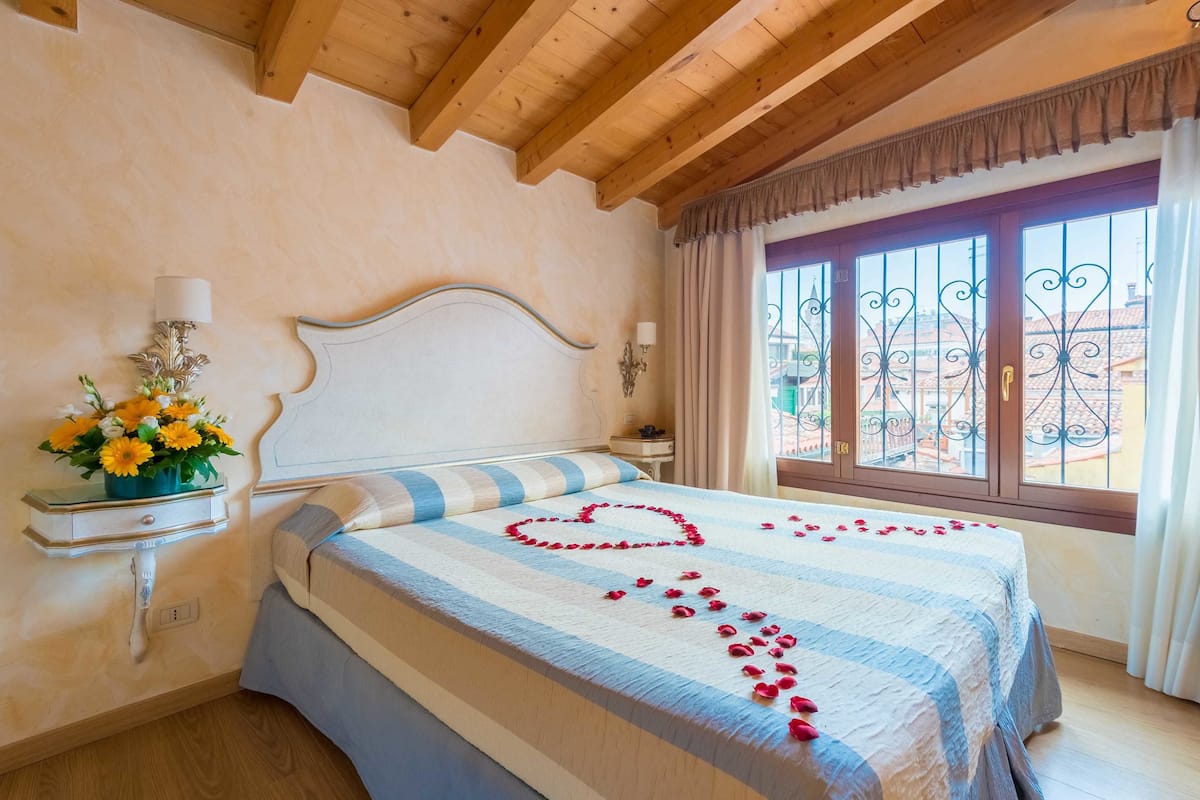 Um quarto com uma cama, mesas de cabeceira, um criado-mudo, uma janela com grades de ferro decorativas e vista para prédios lá fora.