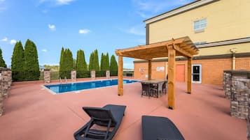 Uma área de piscina com espreguiçadeiras, uma pérgula e um edifício de tijolos ao fundo.