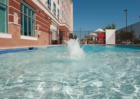 Piscina com uma fonte no centro, rodeada por um edifício de tijolos com janelas grandes e uma área cercada com guarda-sóis.