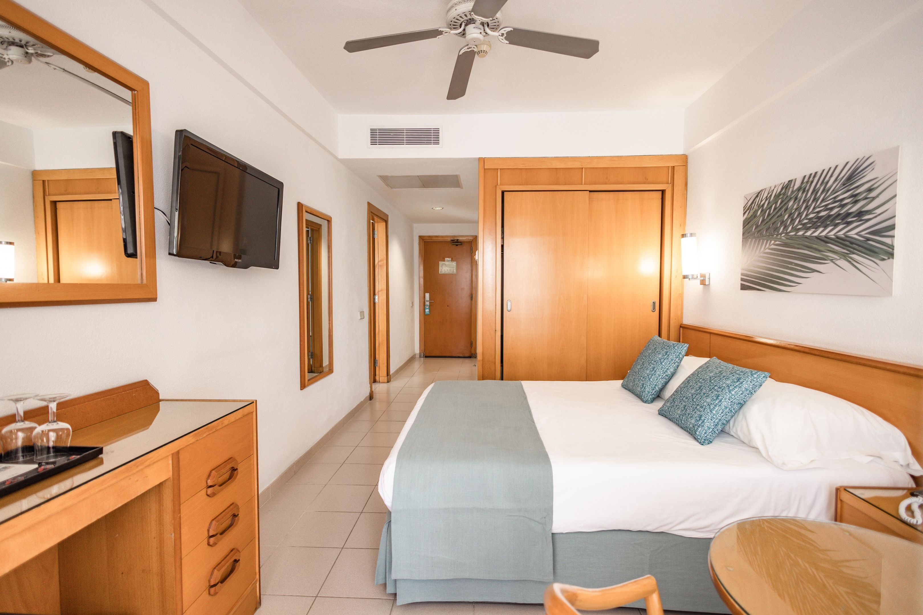 Quarto de hotel com uma cama, escrivaninha com gavetas, televisão, ventoinha no teto e porta que dá para outro cômodo.