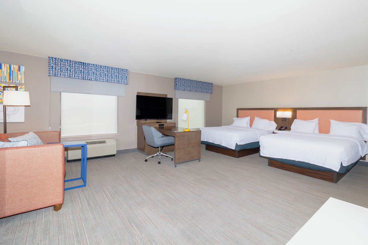 Quarto de hotel com cama, secretária, cadeira, televisão e janela.