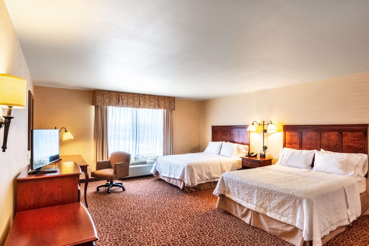 Quarto de hotel com duas camas, uma escrivaninha, uma cadeira, uma televisão e uma janela com cortinas.