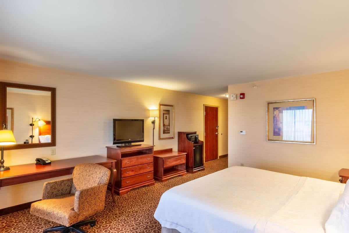 Quarto de hotel com uma cama, uma mesa com abajur, uma cadeira, televisão, guarda-roupa e uma porta.