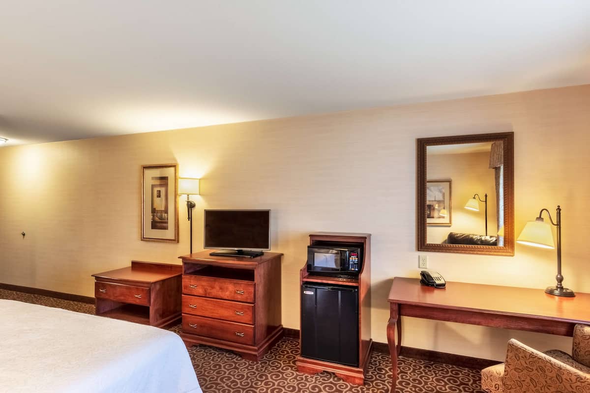 Quarto de hotel com cama, cómoda de madeira, escrivaninha com telefone, micro-ondas, espelho e luminária.
