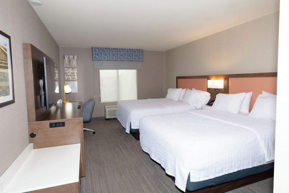 Quarto de hotel com duas camas, uma escrivaninha, uma cadeira, uma janela e obras de arte nas paredes.