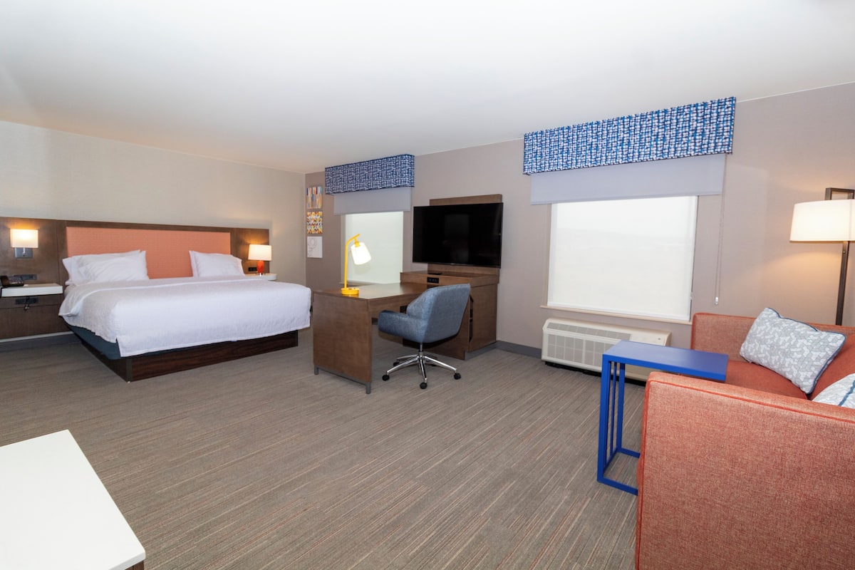 Quarto de hotel com cama, escrivaninha, cadeira, televisão e sofá.