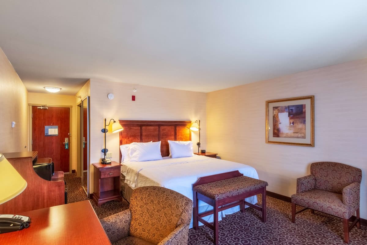 Quarto de hotel com cama, duas poltronas, uma escrivaninha com luminária e uma mesinha.