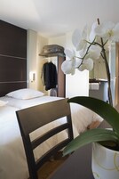 Quarto de hotel com uma cama, uma cadeira, uma orquídea em vaso, um cabideiro suspenso e um banheiro visível através de uma porta aberta.