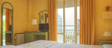 Quarto de hotel com cama, televisão sobre uma mesa, varanda com cortinas e vista para o mar.