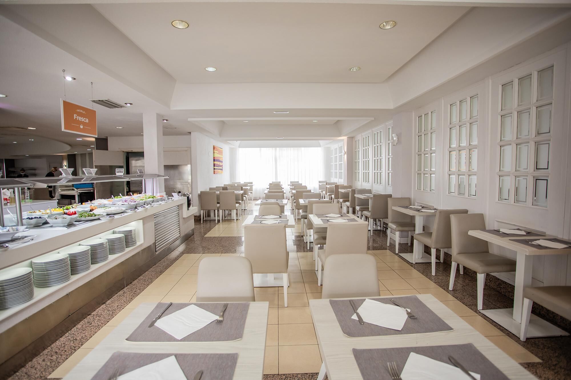 Uma ampla área de refeições com várias mesas e cadeiras, um balcão de buffet com diversos alimentos e um interior claro e bem iluminado.