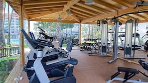 Uma área de ginásio coberta com equipamentos de exercício, incluindo esteiras e máquinas de musculação, sob um telhado de madeira com grandes janelas.