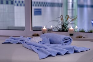 Uma toalha azul bem arrumada e uma concha sobre uma superfície branca, com duas velas acesas e uma planta ao fundo.