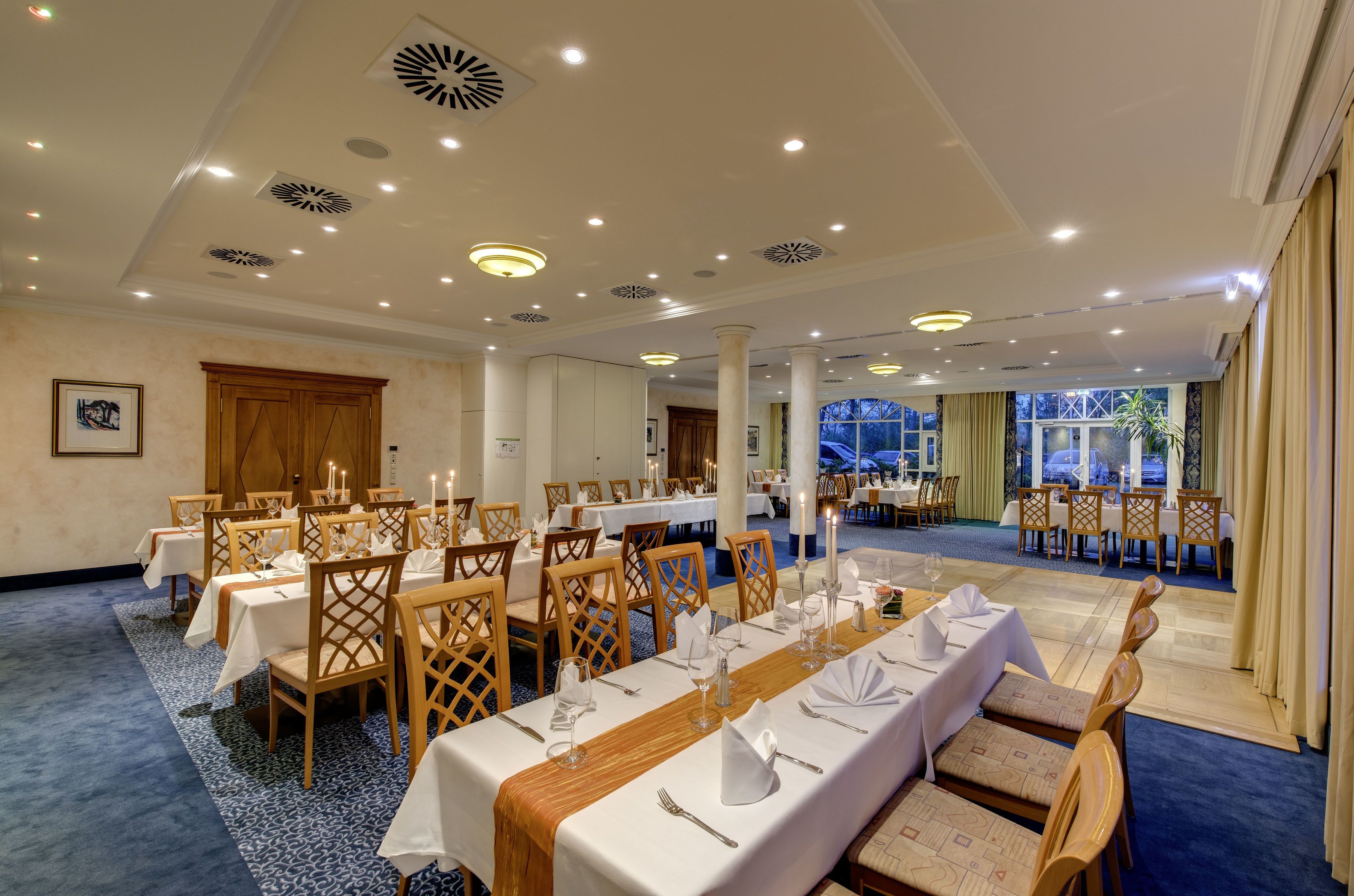 Uma sala de jantar espaçosa com várias mesas preparadas para o serviço, teto alto com iluminação embutida e vista para o exterior.