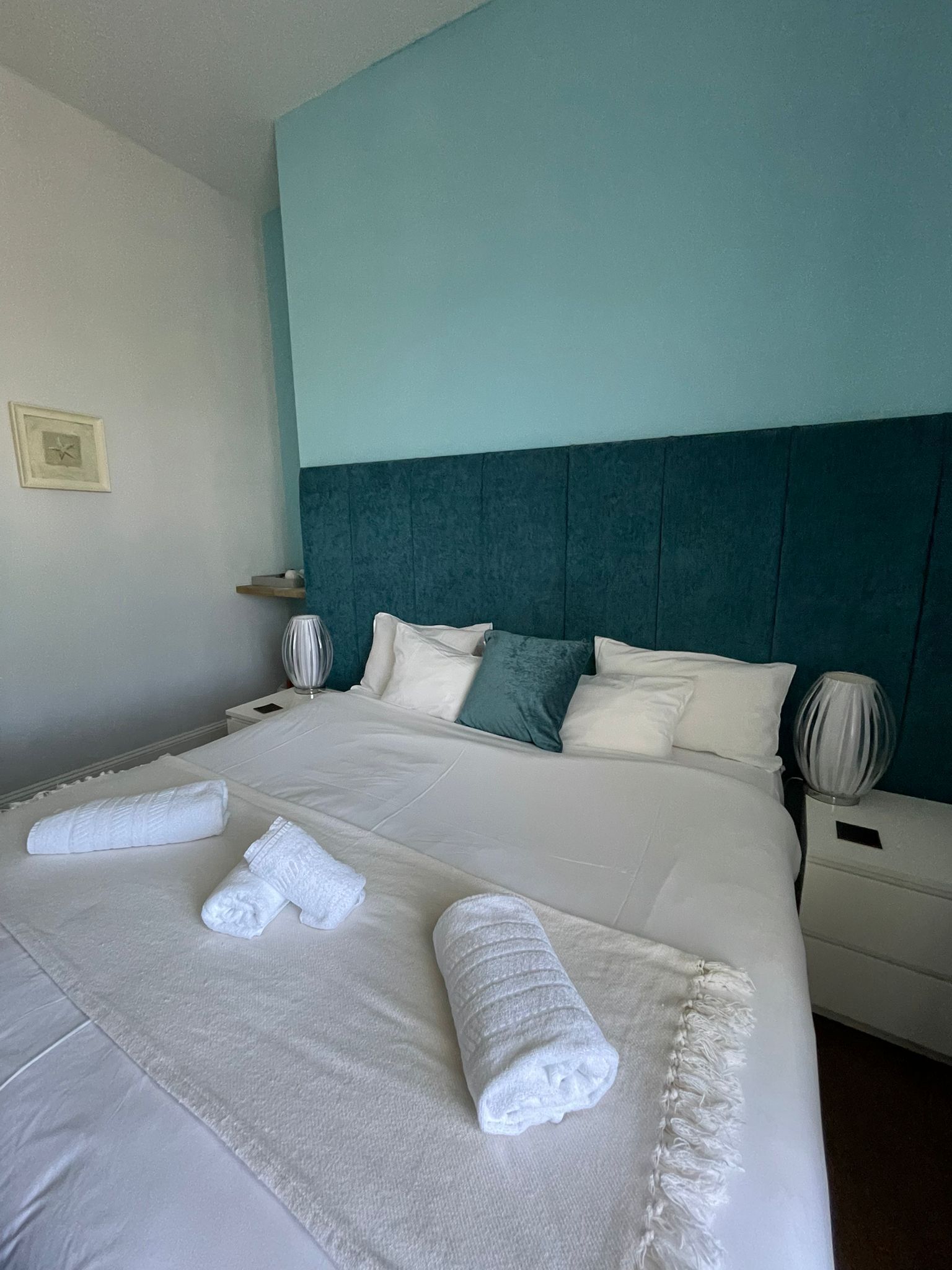 Uma cama bem arrumada com lençóis brancos, cabeceira verde-água e uma luminária de mesa.