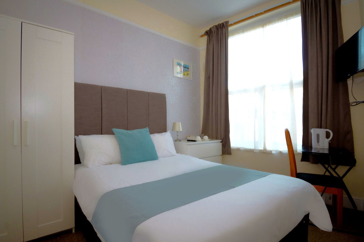 Quarto de hotel com cama, mesa de cabeceira, cadeira, guarda-roupa e televisão na parede.