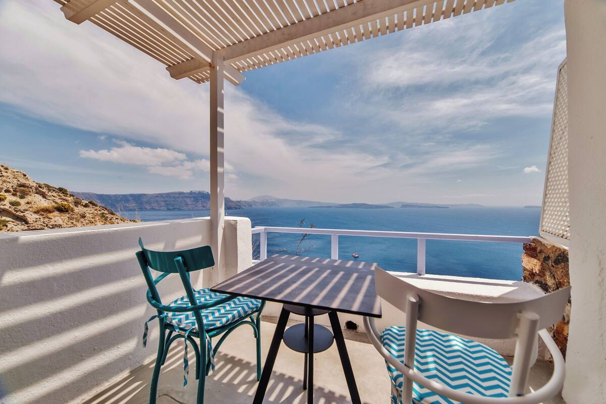 Varanda com mesa e cadeiras, vista para a paisagem costeira, com visão clara do mar e montanhas ao longe.