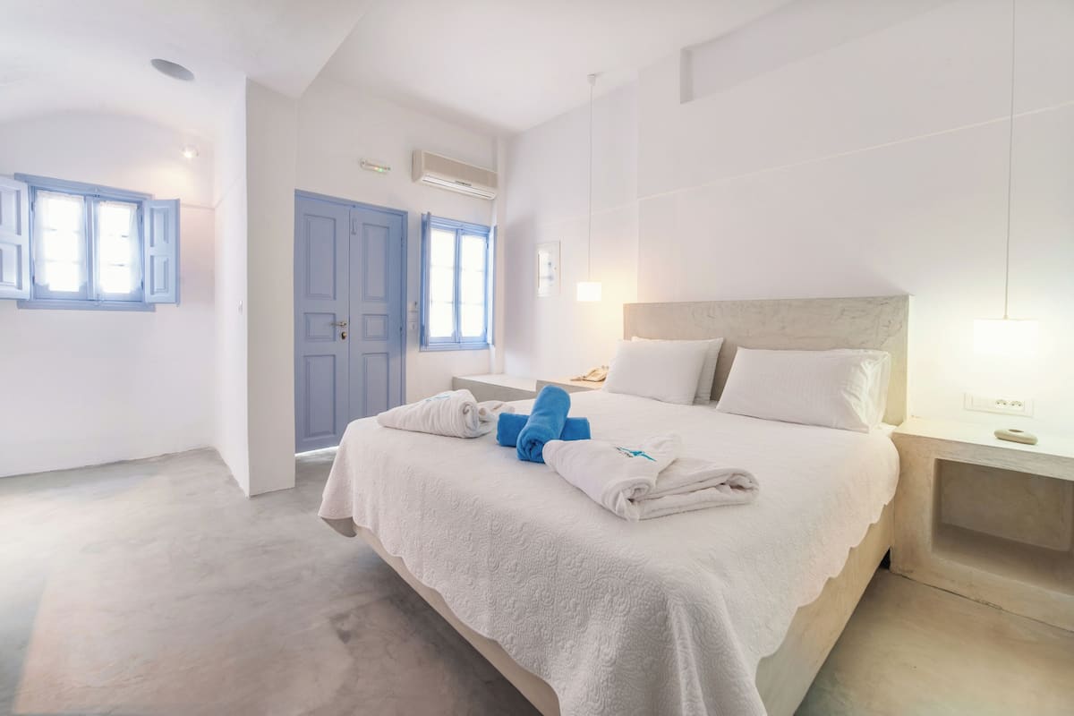 Quarto de hotel com uma cama grande, uma mesa de cabeceira e uma janela com persianas azuis.