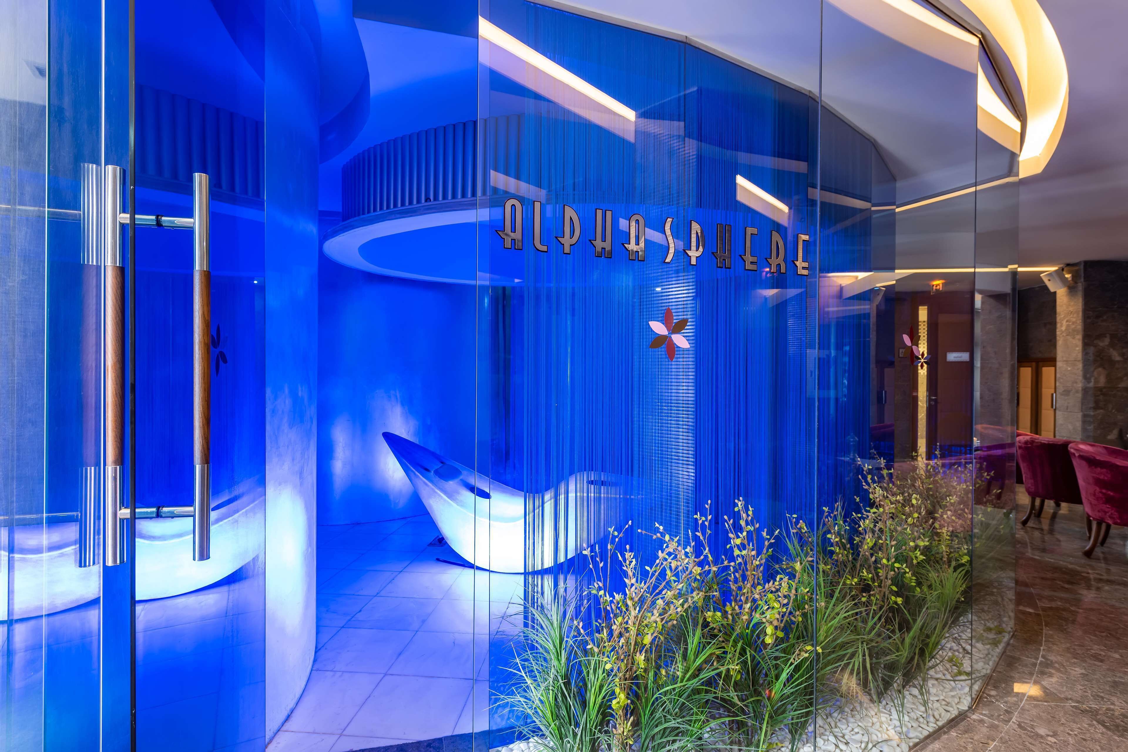 Uma entrada de hotel moderna com fachada de vidro azul, uma recepção visível e uma placa com a inscrição "ALPHA SPREE".