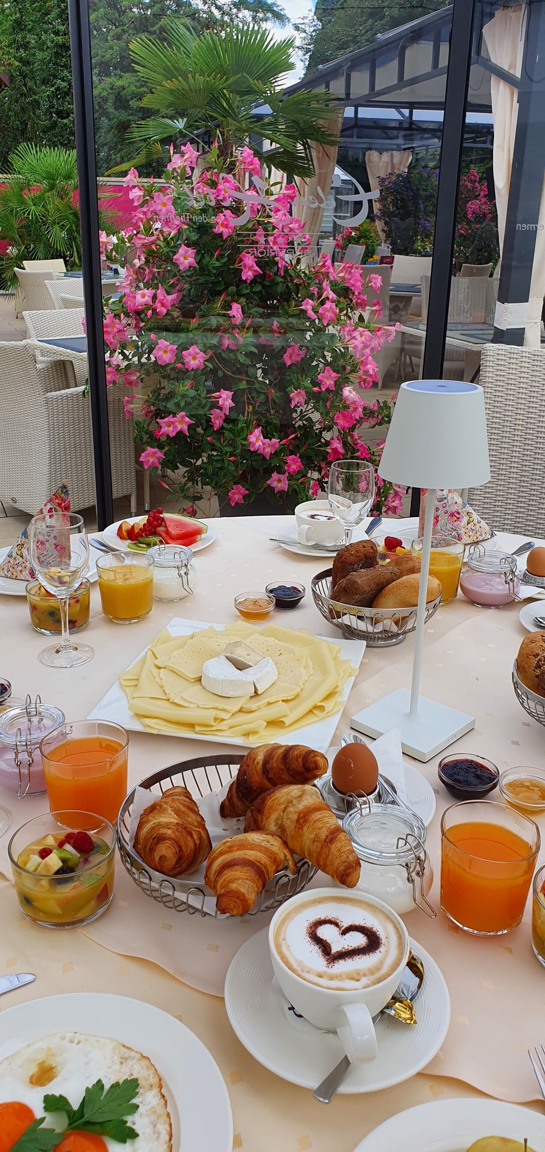 Mesa de café da manhã posta com croissants, waffles e café, rodeada por flores cor-de-rosa e vegetação.