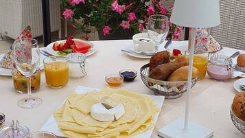 Mesa de café da manhã posta com croissants, waffles e café, rodeada por flores cor-de-rosa e vegetação.