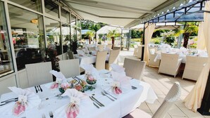 Uma área de refeições ao ar livre com mesas montadas para um evento formal, com toalhas brancas, arranjos florais e cadeiras de vime.