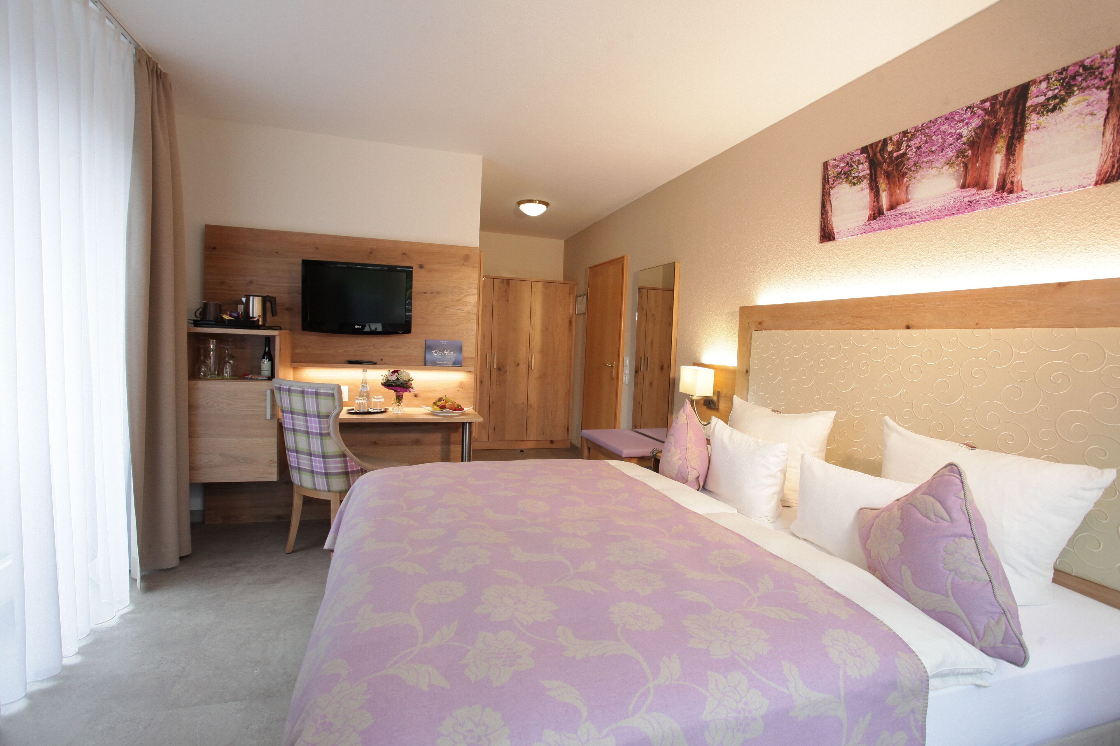 Quarto de hotel com uma cama grande, uma escrivaninha com cadeira, televisão e um quadro na parede.