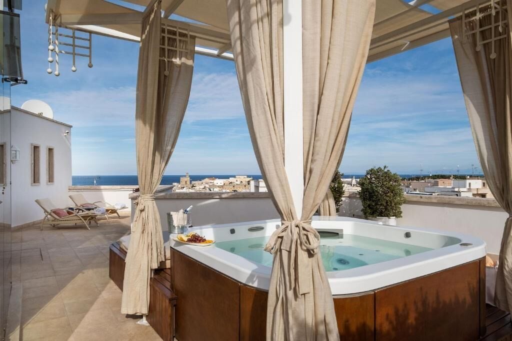 Terraço no telhado com hidromassagem, cortinas bege, cadeiras de descanso e vista para o mar.