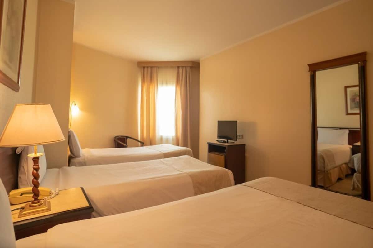 Quarto de hotel com duas camas, uma escrivaninha com luminária, televisão e espelho.